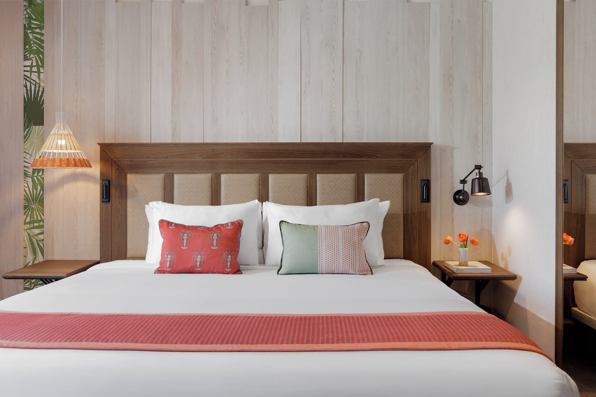 Quarto de hotel com uma cama grande, duas mesas de cabeceira, um criado-mudo, uma luminária na parede e uma cabeceira de madeira.