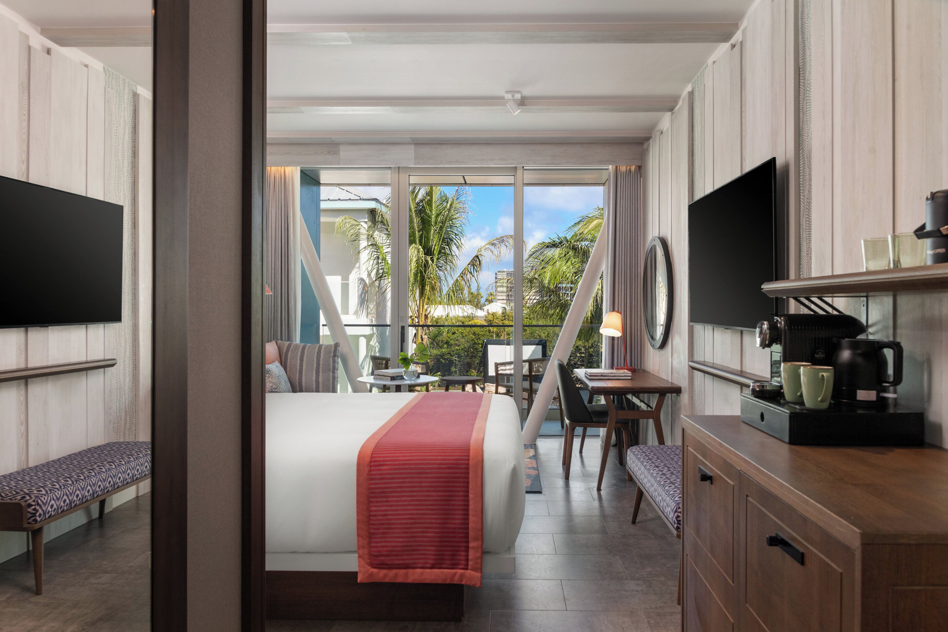 Quarto de hotel com cama, escrivaninha, cadeira, televisão e varanda com vista para palmeiras.