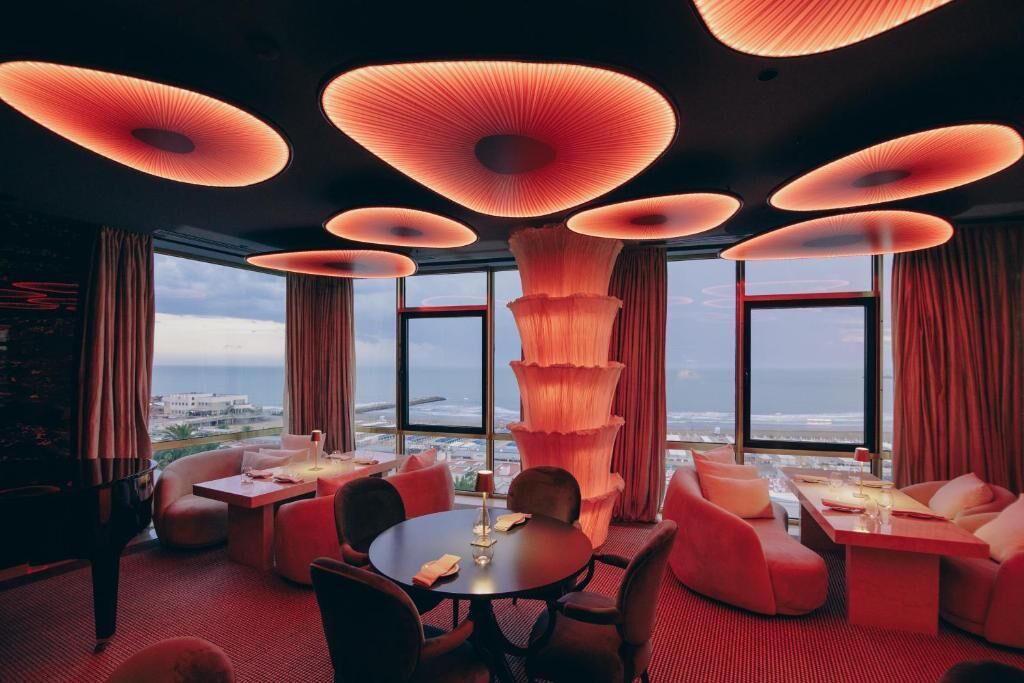Interior moderno com luminárias redondas no teto, área de jantar com mobiliário vermelho e grandes janelas com vista para a cidade.