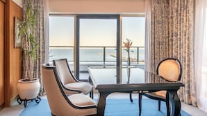 Quarto de hotel com varanda, escrivaninha, duas cadeiras, um vaso de planta e vista para o mar.