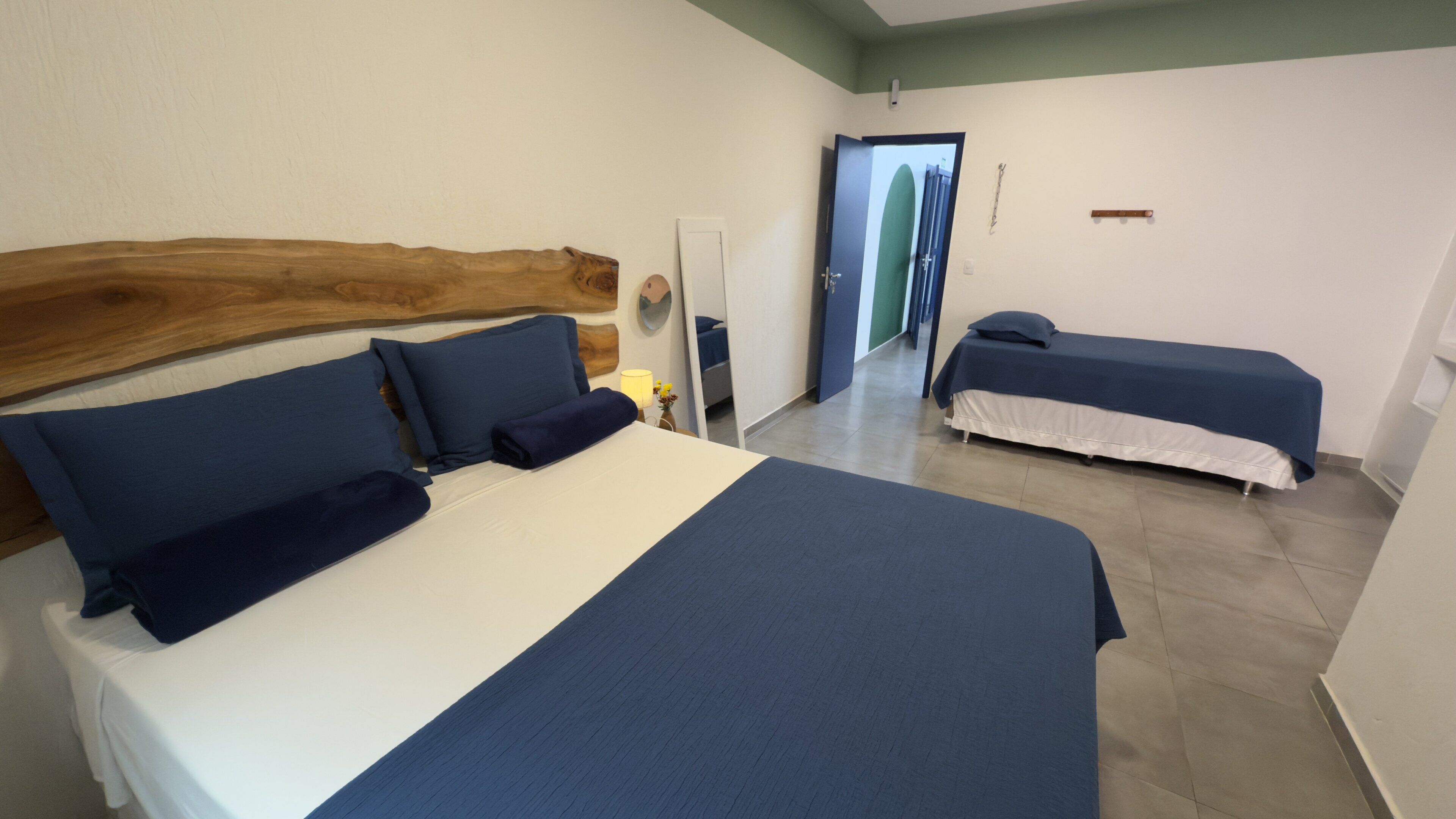 Quarto com uma cama grande, duas camas menores, cabeceira de madeira, mesa de cabeceira com abajur e porta que d á para outro cômodo.