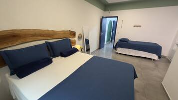 Quarto com uma cama grande, duas camas menores, cabeceira de madeira, mesa de cabeceira com abajur e porta que dá para outro cômodo.