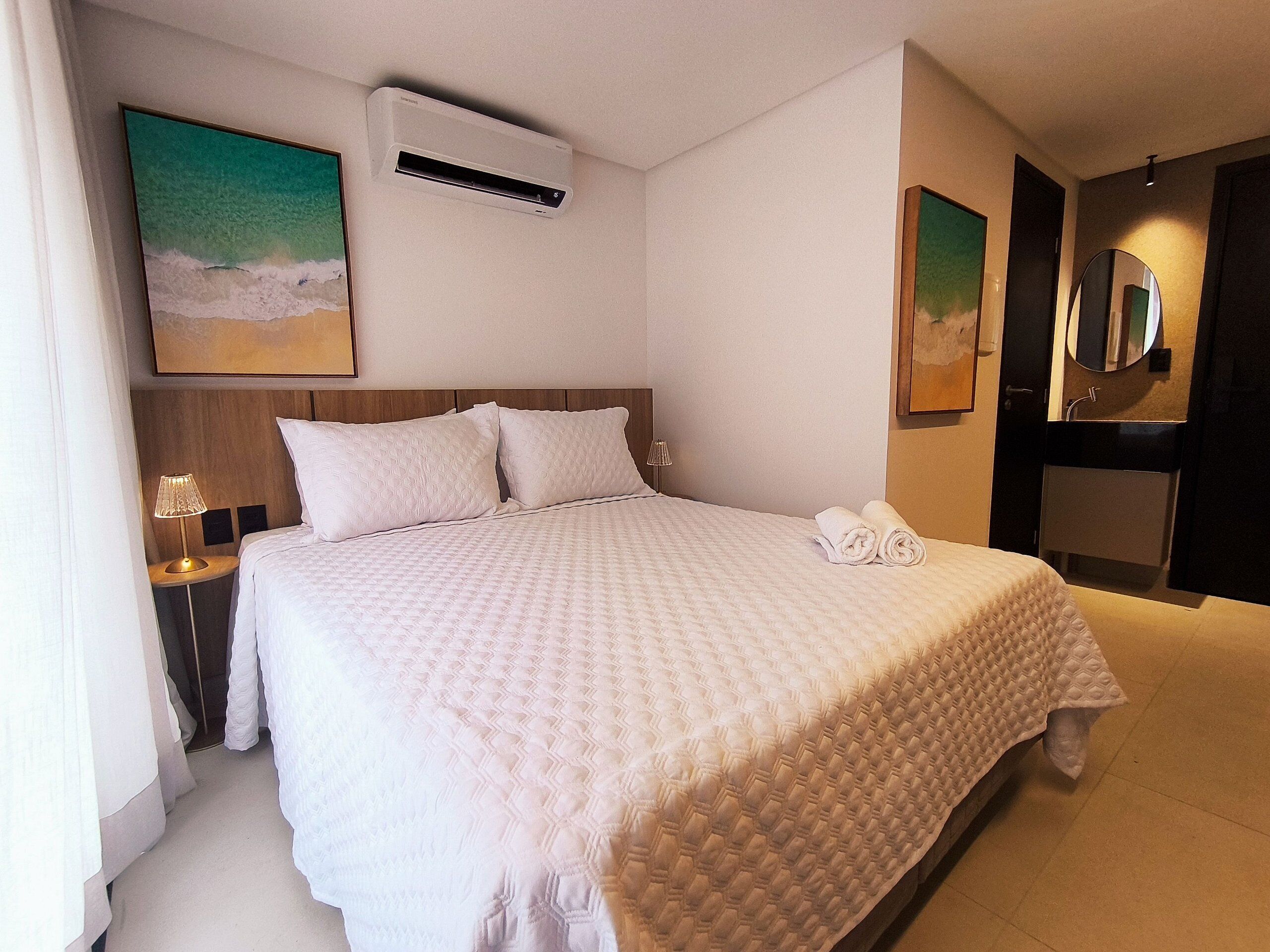Quarto de hotel com uma cama grande, ar condicionado fixado na parede, pintura de uma praia, espelho e banheiro com pia e espelho.