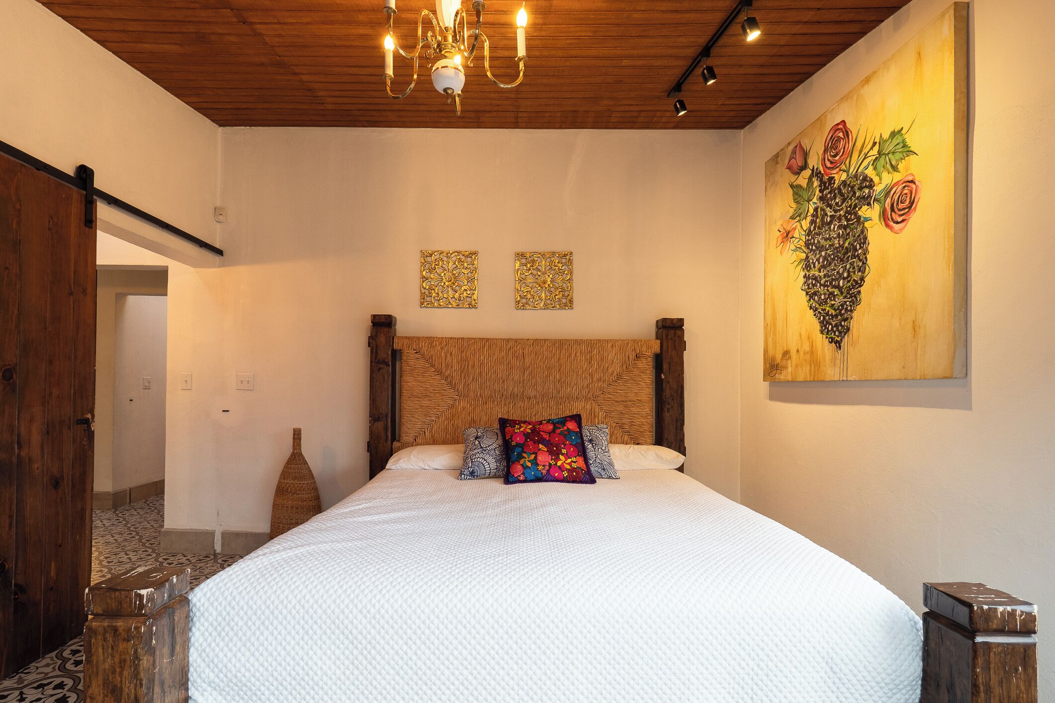 Quarto com teto de madeira, cama com roupa de cama branca, duas peças decorativas na parede e um quadro de uvas e rosas.