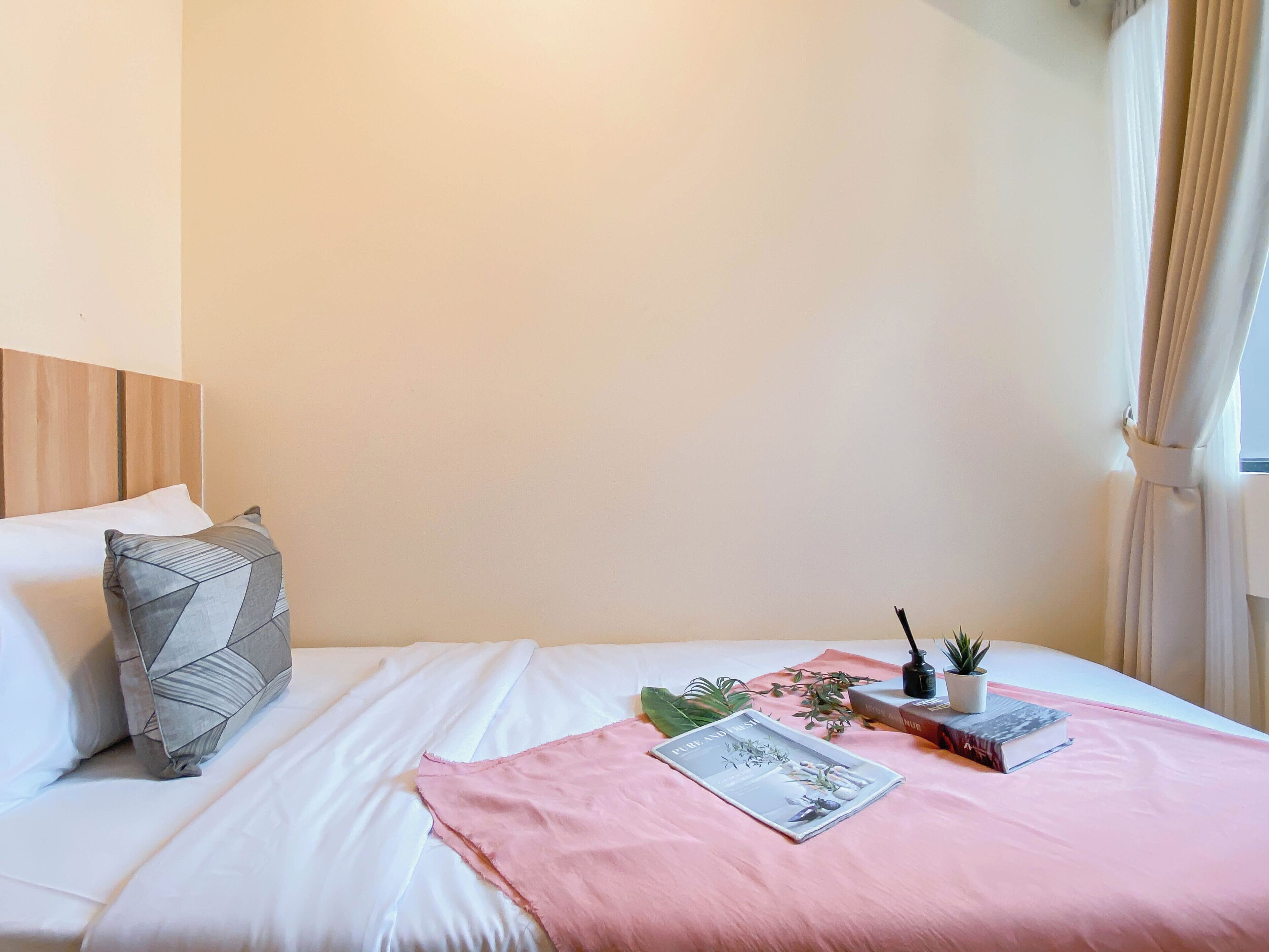 Uma cama bem arrumada com um cobertor rosa, uma almofada decorativa, uma pequena planta em vaso e um livro sobre a cama.