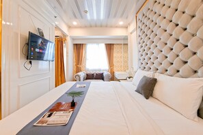 Quarto de hotel com uma cama grande, televisão de tela plana fixada na parede, janela com cortinas e cabeceira estofada.