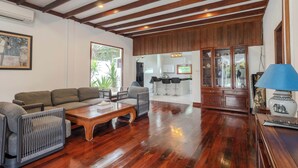 Ampla sala de estar com teto de madeira, um sofá, uma mesa de centro e área de cozinha visível por uma porta aberta.