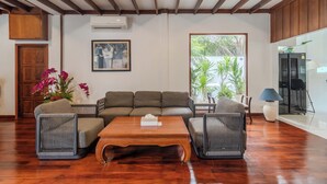 Uma sala de estar moderna com teto de madeira, um sofá, duas poltronas, uma mesa de centro e uma janela grande com vista para vegetação.