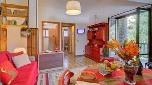 Uma sala de estar acolhedora com um sofá vermelho, um armário de madeira, uma mesa de jantar com um vaso de folhas e uma televisão ao fundo.