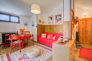 Um quarto aconchegante com um sofá vermelho, paredes de painel de madeira, uma mesa de jantar com cadeiras, um lustre pendurado e um pequeno armário de madeira.