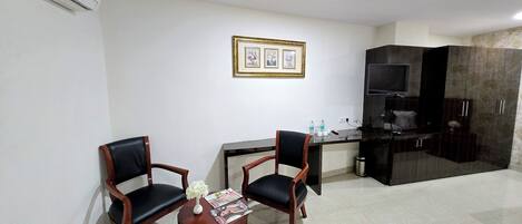 Quarto pequeno e bem iluminado com duas cadeiras de madeira, uma mesinha com um vaso e uma revista, um quadro na parede e um armário escuro com uma televisão.