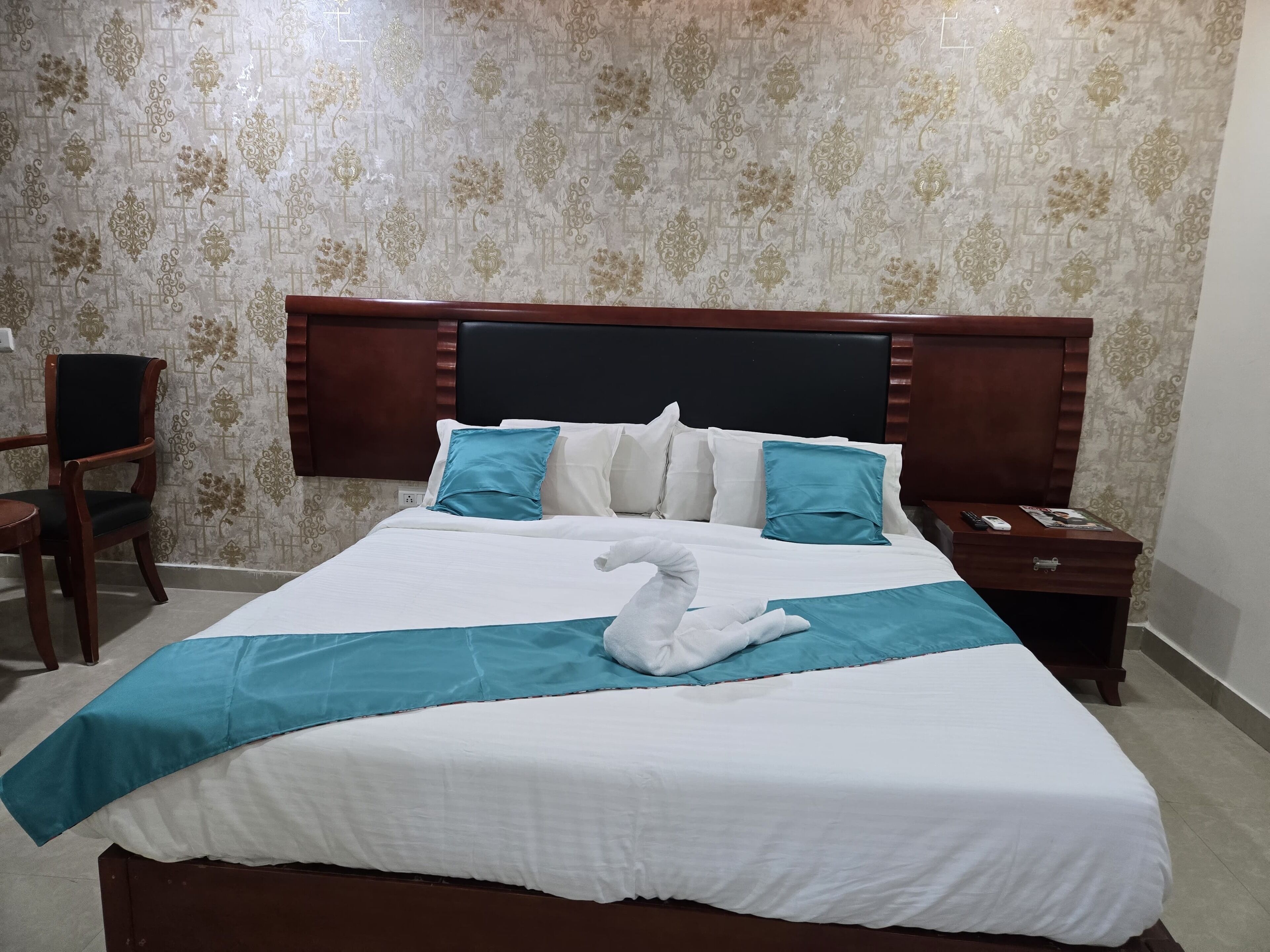 Cama bem arrumada com colcha branca e azul-turquesa, almofada azul-turquesa combinando e toalha branca dobrada sobre a cama.