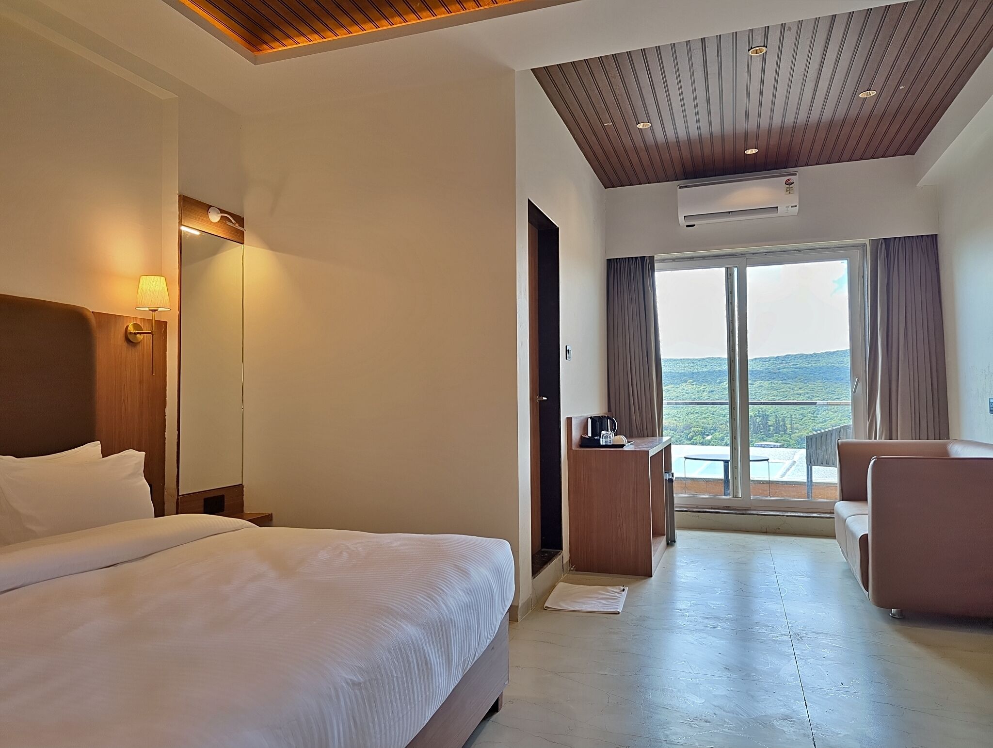 Quarto de hotel com cama, escrivaninha, cadeira e uma grande janela com vista para o exterior.