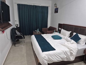 Quarto de hotel com cama, cadeira, escrivaninha, televisão e janela com cortinas.