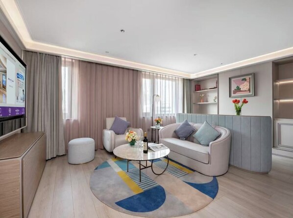 Quarto de hotel moderno com sofá, poltrona, banqueta e mesa de centro redonda com controle remoto.