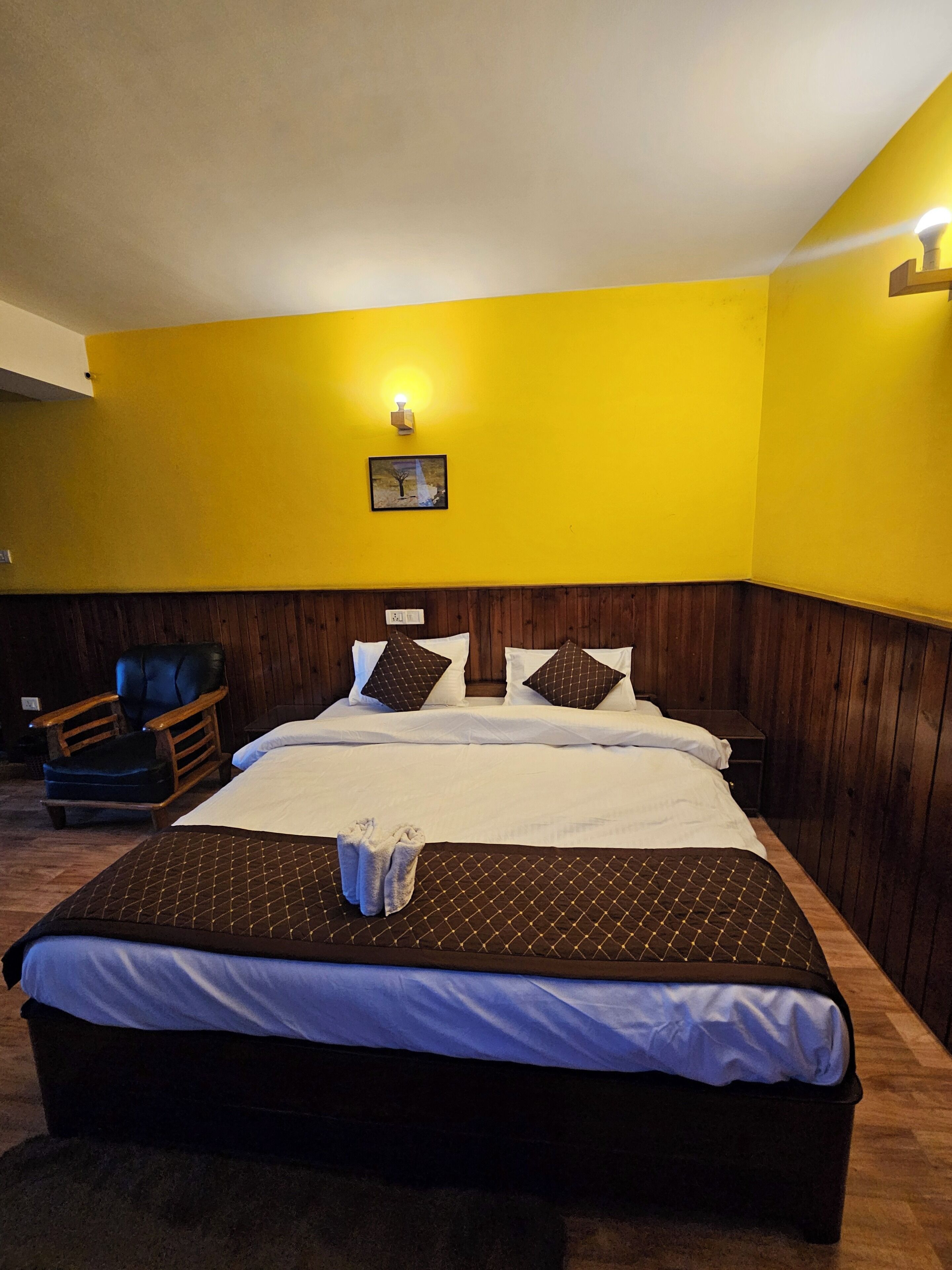 Quarto de hotel com uma cama grande, cabeceira de madeira, paredes amarelas e uma luminária fixada na parede.