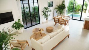 Uma sala de estar moderna com sofá, mesa de jantar e cadeiras, cercada por amplas janelas e plantas em vasos.