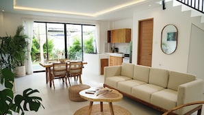 Uma sala de estar moderna com sofá, mesa de jantar, cadeiras e mesa de centro. Há uma área de cozinha com armários de madeira e micro-ondas. O quarto tem janelas amplas com vista para o verde.