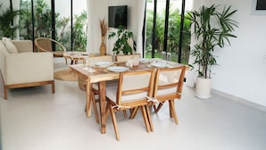 Uma área de refeições com mesa e cadeiras de madeira, cercada por plantas em vasos e grandes janelas.