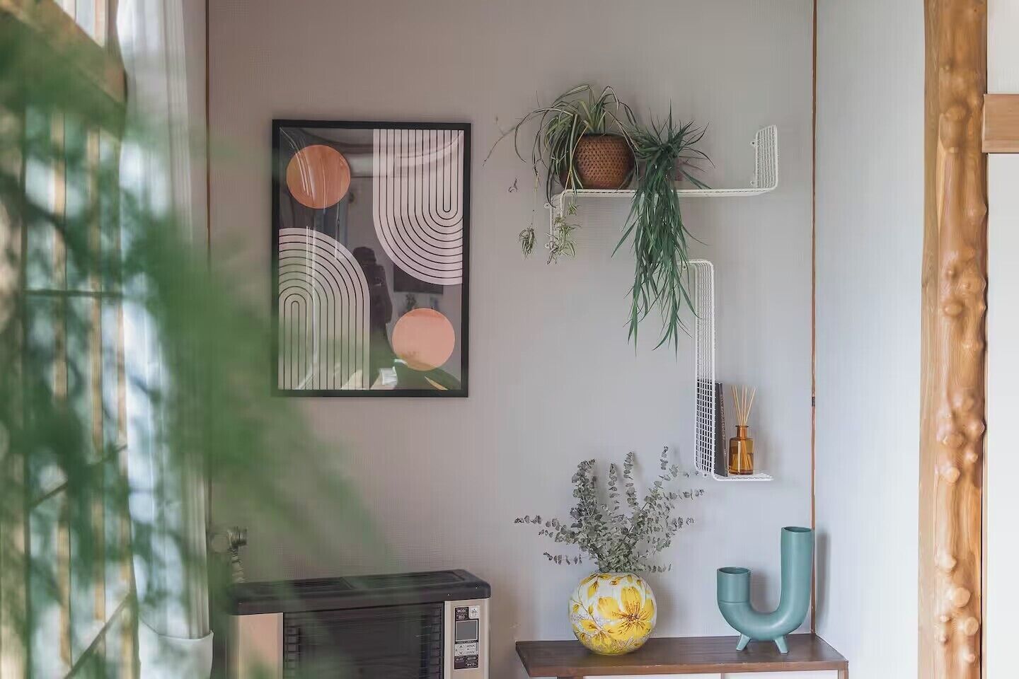 Quarto com decoração moderna, incluindo uma obra de arte abstrata emoldurada, um vaso de plantas, uma pequena prateleira com um frasco e difusor de vagem, e um vaso com flores.