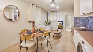 Uma cozinha moderna com máquina de lavar embutida, um espaço de refeições com mesa redonda e cadeiras, e uma sala de estar com sofá e um espelho grande.