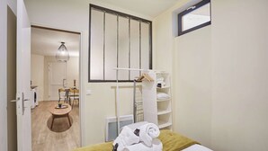Quarto de hotel com uma cama, uma mesinha, uma cadeira, uma janela, um porta-toalhas e um banheiro visível através de uma porta aberta.