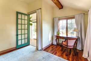 Quarto com piso de madeira, uma escrivaninha com cadeira, janela com cortinas e porta com moldura verde.
