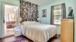Quarto com cama, mesa de cabeceira, cômoda, janela com cortinas e porta que dá para uma varanda.