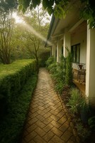 Uma trilha pavimentada conduz a um edifício com varanda coberta, rodeado por vegetação exuberante e árvores.
