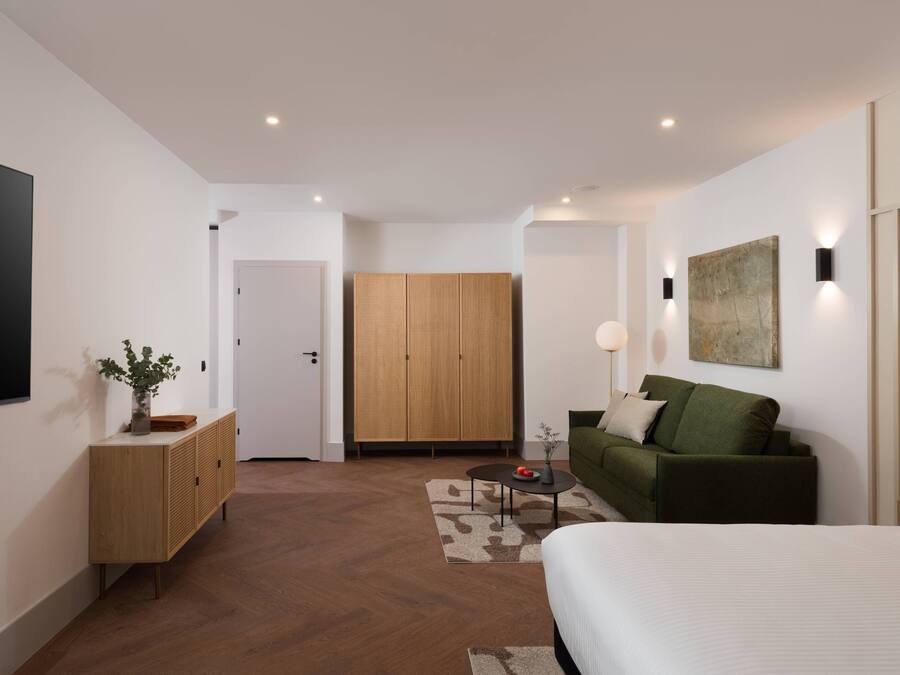 Quarto de hotel moderno com uma cama grande, um sofá verde, um guarda-roupa de madeira e uma televisão de tela plana fixada na parede.