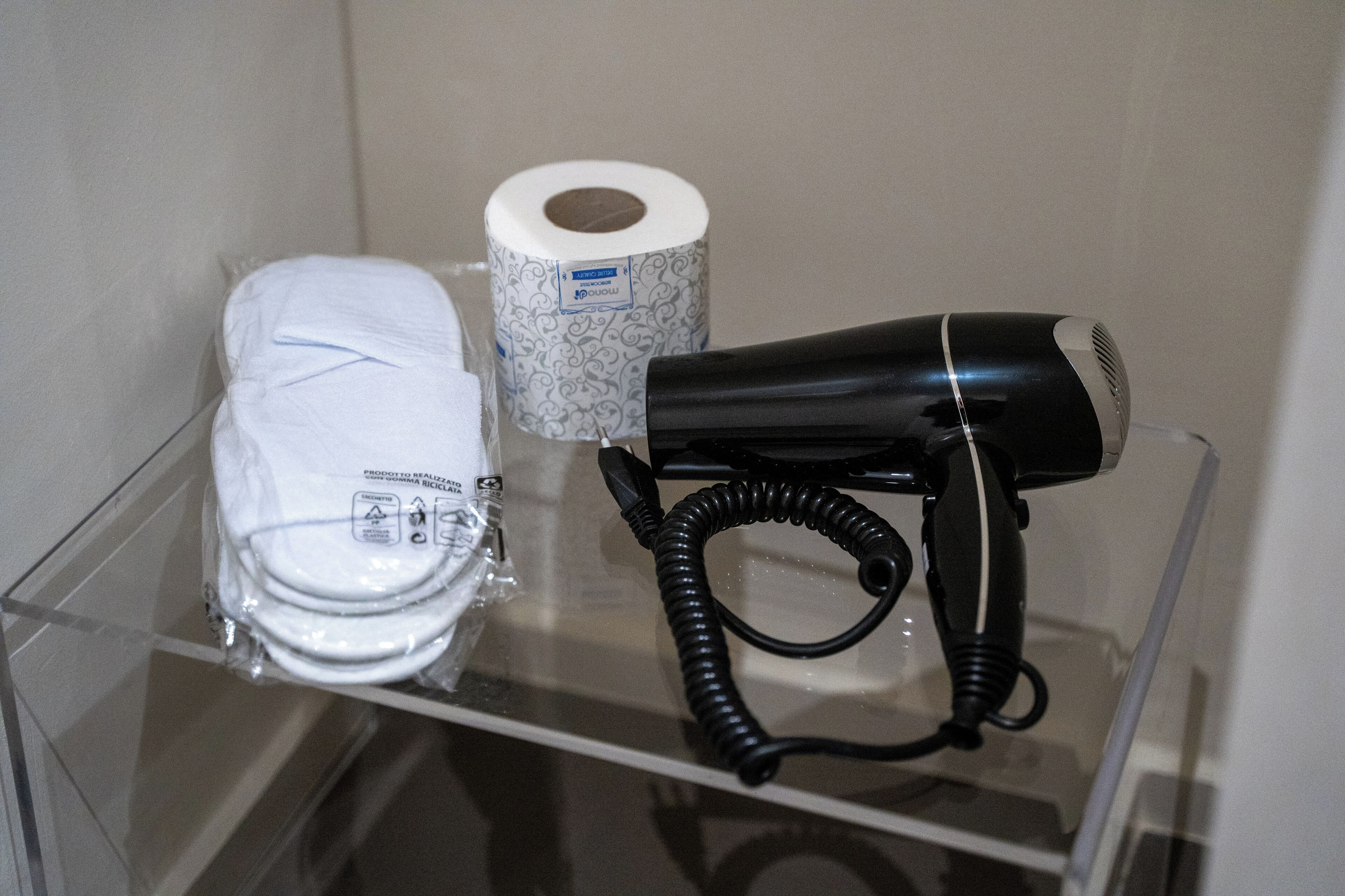 Um secador de cabelo, um rolo de papel higiénico e uma embalagem de guardanapos brancos numa prateleira de vidro transparente.