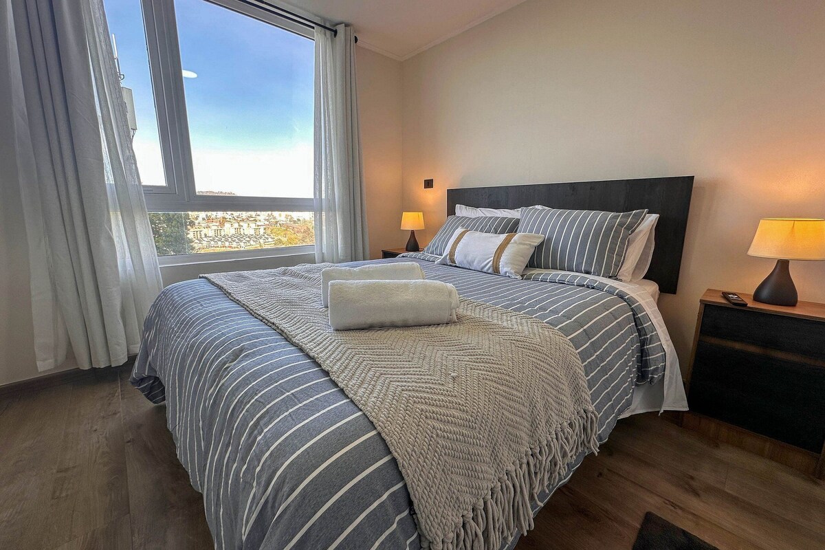 Quarto de hotel com uma cama grande, mesa de cabeceira com abajur e vista da cidade pela janela.