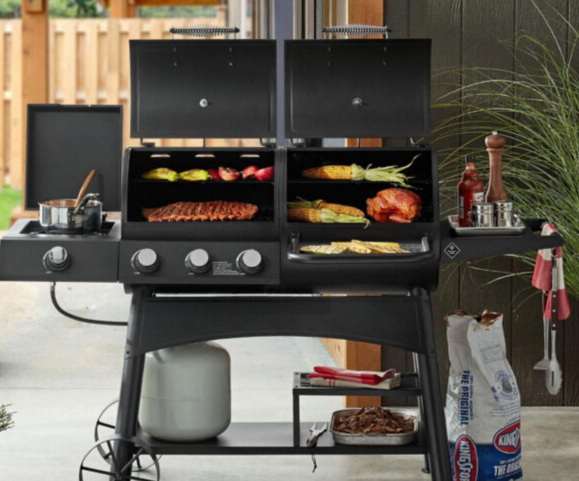 Uma churrasqueira preta com várias zonas de cozimento, uma mesa lateral com acompanhamentos e um saco de carvão por perto.