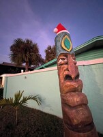 Um totém de madeira com rosto entalhado e gorro de Papai Noel, diante de um edifício com telhado verde e palmeiras ao fundo.