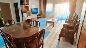 Sala de estar com uma mesa de madeira redonda, cadeiras combinando, televisão, prateleiras e uma janela grande com cortinas.