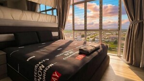 Quarto moderno com uma cama grande, cabeceira e vista da cidade através de janelas amplas.