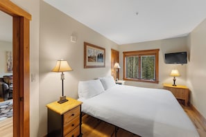 Um quarto com uma cama grande, duas mesas de cabeceira com luminárias, uma TV fixada na parede e uma janela com vista para árvores.