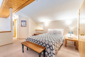 Quarto com cama, mesa de cabeceira com abajur, um banco e um quadro na parede.