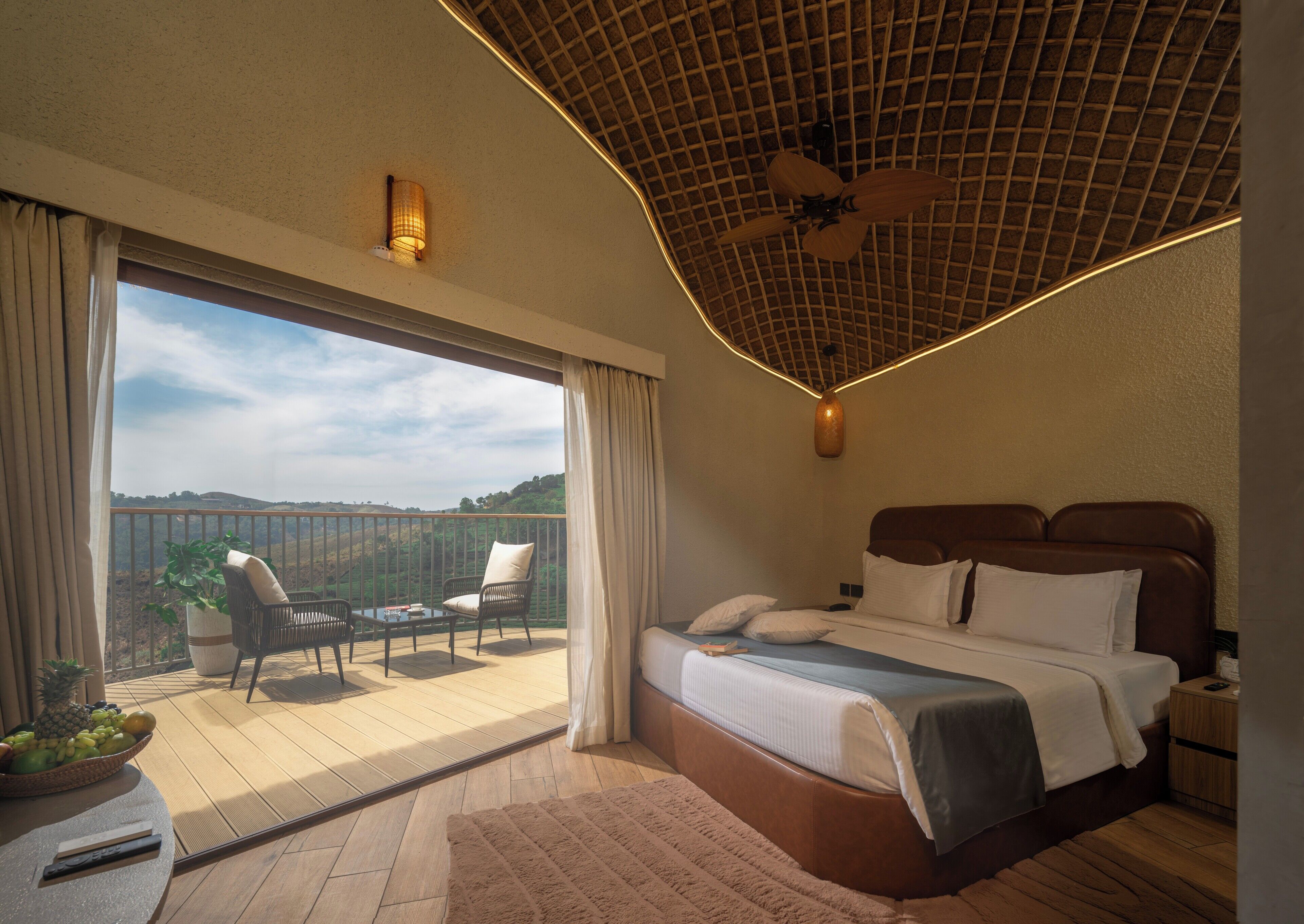 Quarto com uma cama grande, um ventilador de teto, uma varanda com vista para as colinas e uma mesa com uma planta e frutas.