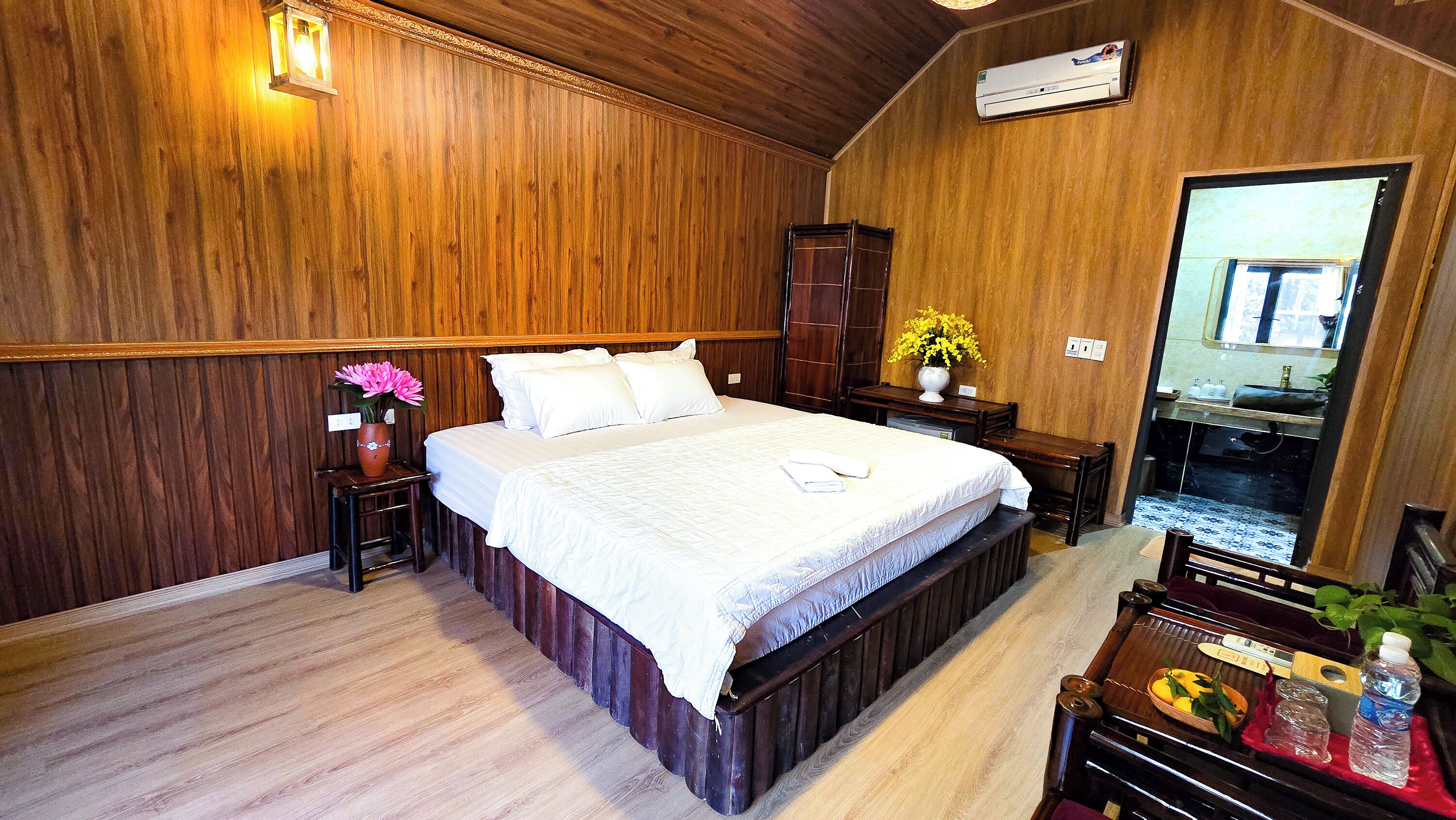 Quarto com painéis de madeira, uma cama grande, mesa de cabeceira, cômoda e espelho.