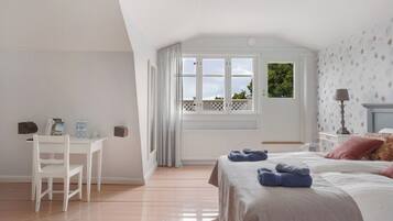 Quarto com cama, escrivaninha e cadeira. Tem uma janela com cortinas, um radiador e uma luminária em uma mesinha de cabeceira.