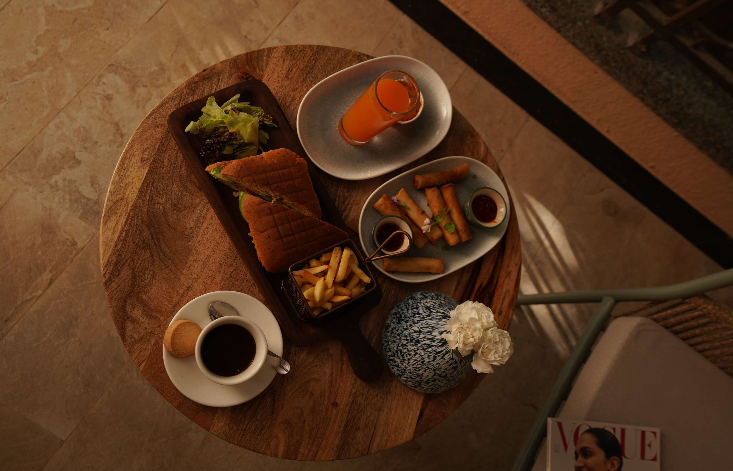 Uma mesa de madeira redonda com diversos alimentos, incluindo um sanduíche, batatas fritas, rolinhos primavera e um copo de suco de laranja. Também há duas xícaras de café e um vaso decorativo.