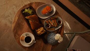 Uma mesa de madeira redonda com diversos alimentos, incluindo um sanduíche, batatas fritas, rolinhos primavera e um copo de suco de laranja. Também há duas xícaras de café e um vaso decorativo.