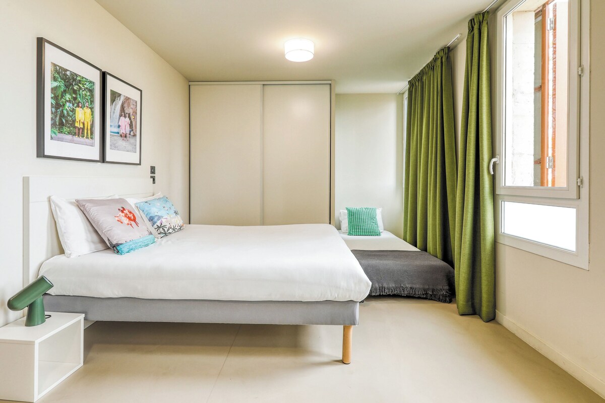 Quarto com cama, mesa de cabeceira, guarda-roupa, janela com cortinas e duas imagens emolduradas na parede.