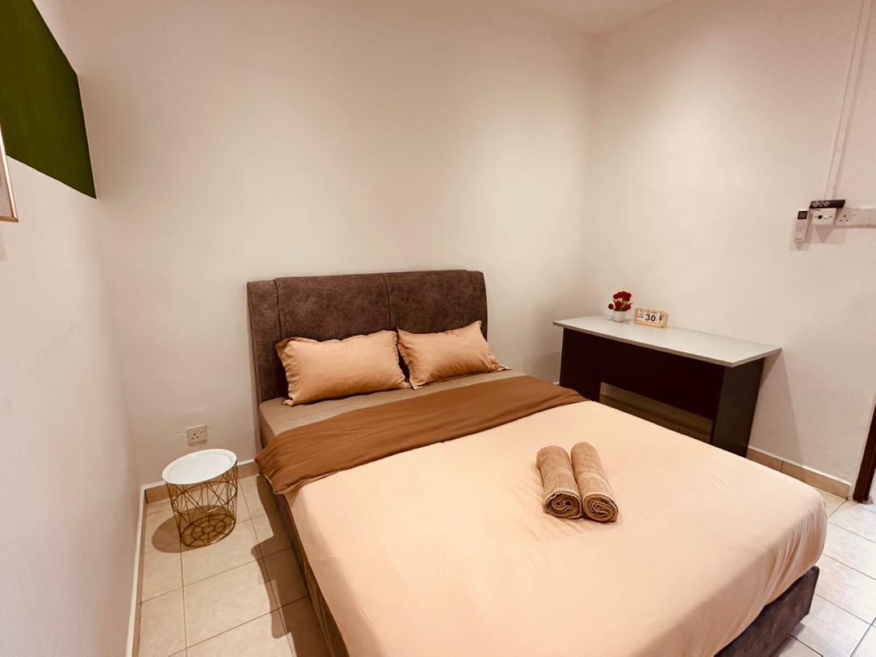 Quarto de hotel com cama, mesa de cabeceira, pequena mesa redonda com base em fio dourado e tomada na parede.