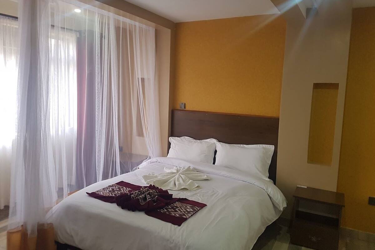 Quarto de hotel com cama, roupa de cama branca, cabeceira de madeira, mesa de cabeceira e cortinas finas.