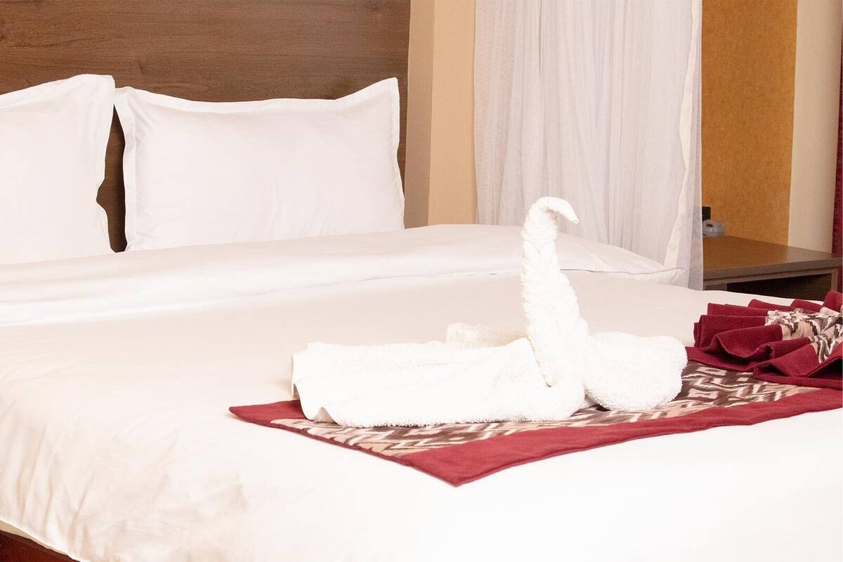 Cama bem arrumada com lençóis brancos, manta com padrão vermelho e bege, e toalha branca dobrada em formato de cisne.