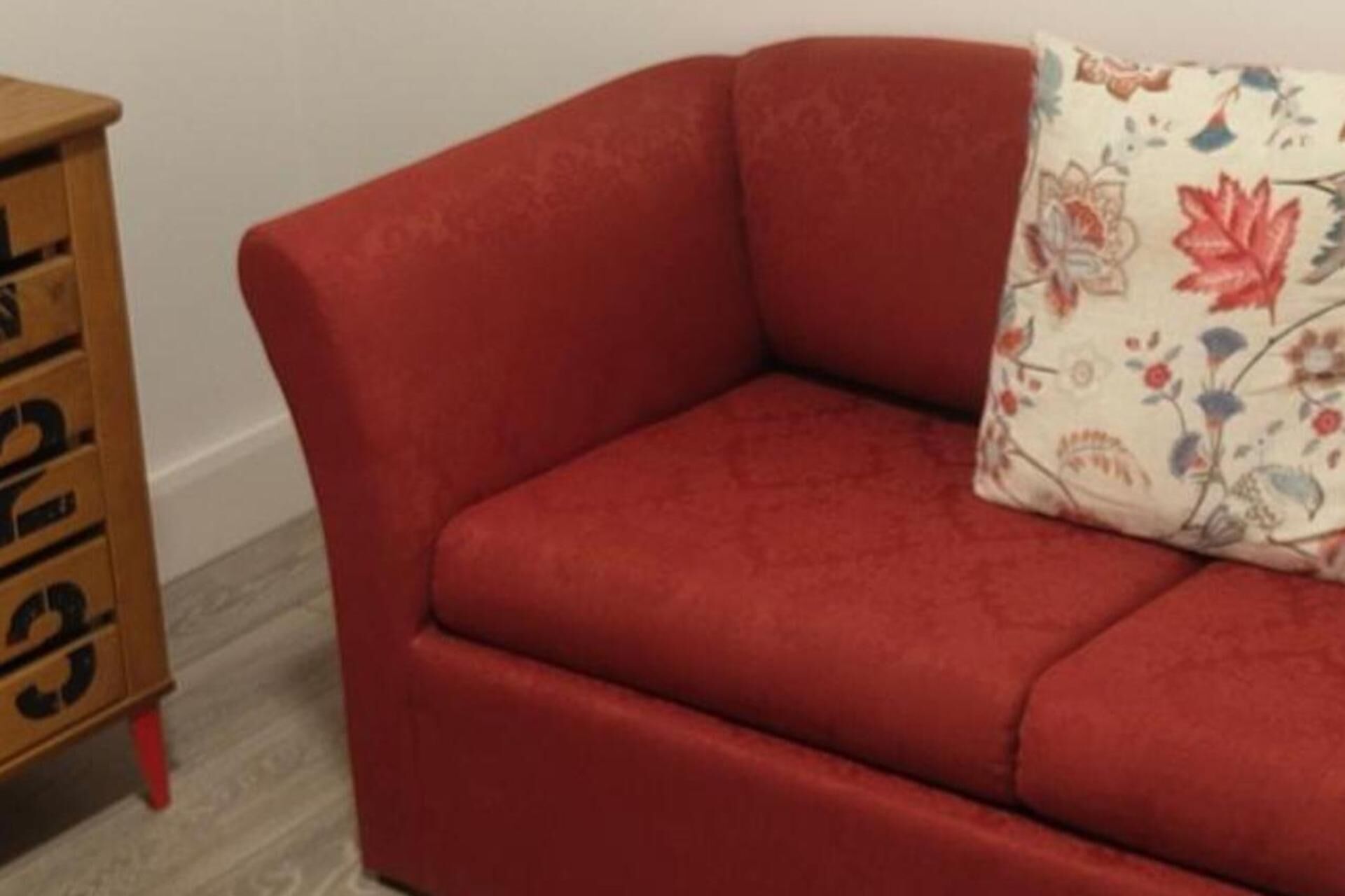 Um sofá vermelho com uma almofada florida e uma mesinha de cabeceira de madeira com gavetas.