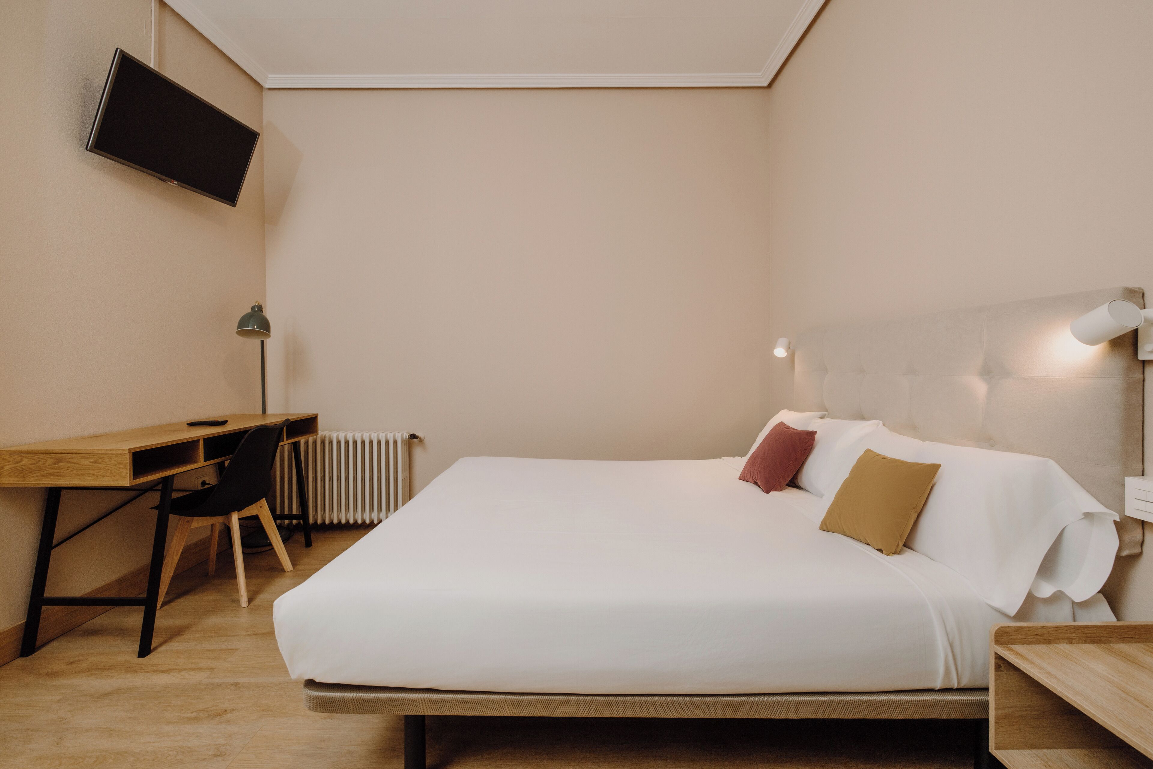 Quarto de hotel com cama, escrivaninha, cadeira, televisão na parede e abajur.