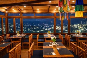 Restaurante com mobiliário de madeira e grandes janelas, oferecendo vista noturna da cidade.